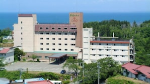 Edifício com vários andares, estacionamento em frente, rodeado por vegetação e vista para o mar.