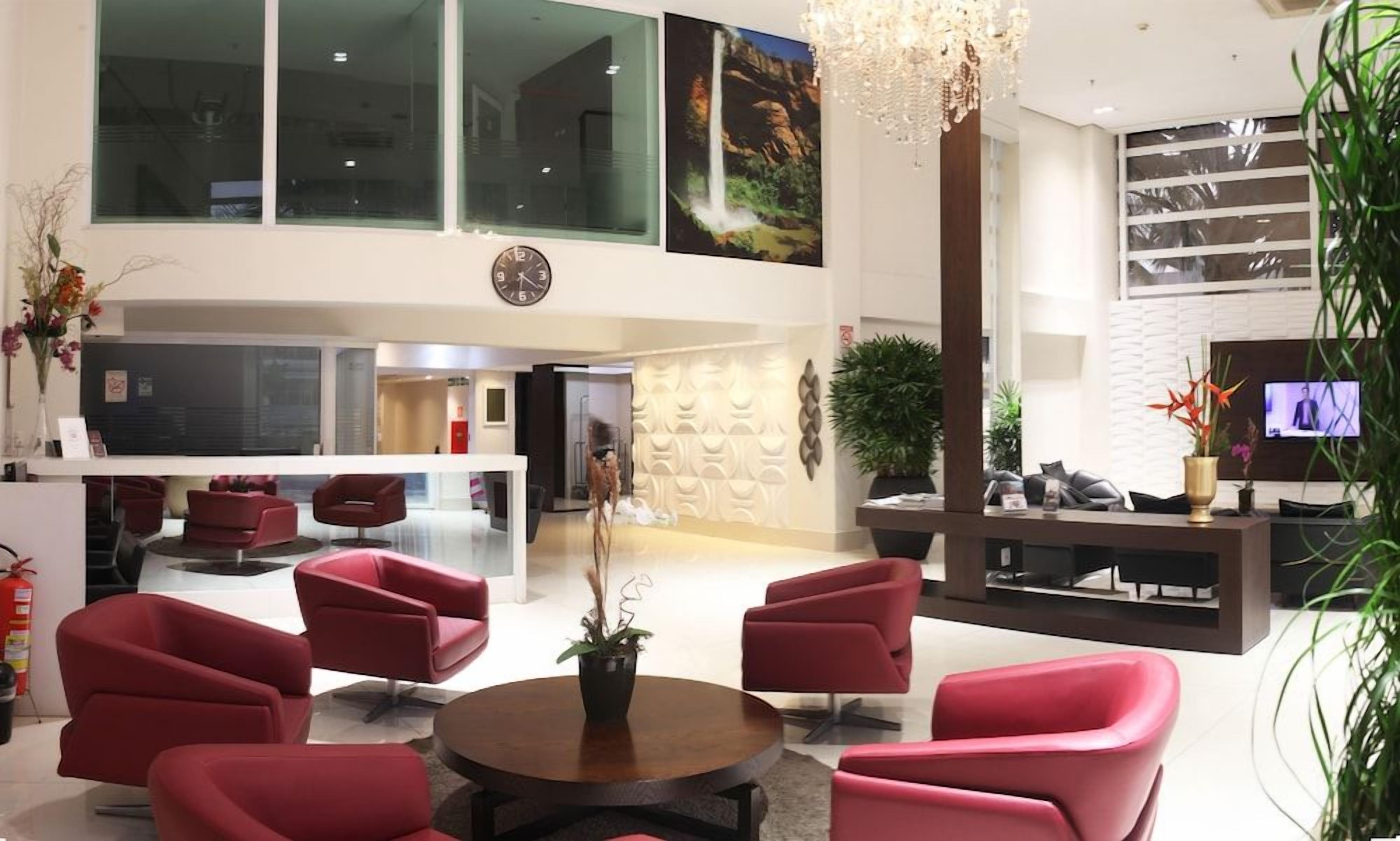 Um hall de hotel moderno com assentos vermelhos, um balcão de recepção, uma janela ampla e uma televisão.