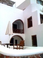 Um edifício branco com arcadas e janelas, área de estar ao ar livre com mesa e cadeiras, e um guarda-sol branco.