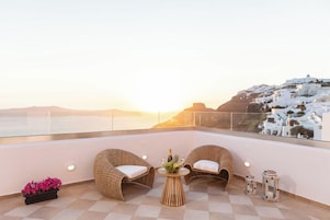 Varanda com cadeiras de vime, uma pequena mesa com flores e vista para edifícios brancos ao pôr do sol.