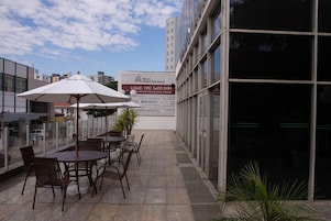 Uma área externa com mesas e cadeiras, um guarda-sol grande e um prédio com uma placa.