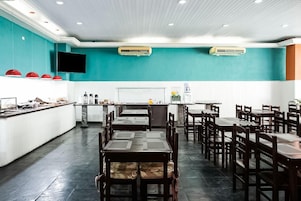 Restaurante com um balcão comprido, várias mesas e cadeiras. Há luminárias no teto e um aparelho de ar condicionado.