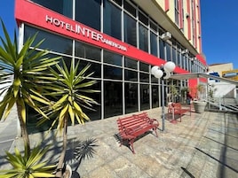 Fachada de hotel com placa vermelha, grandes janelas e área de estar externa.