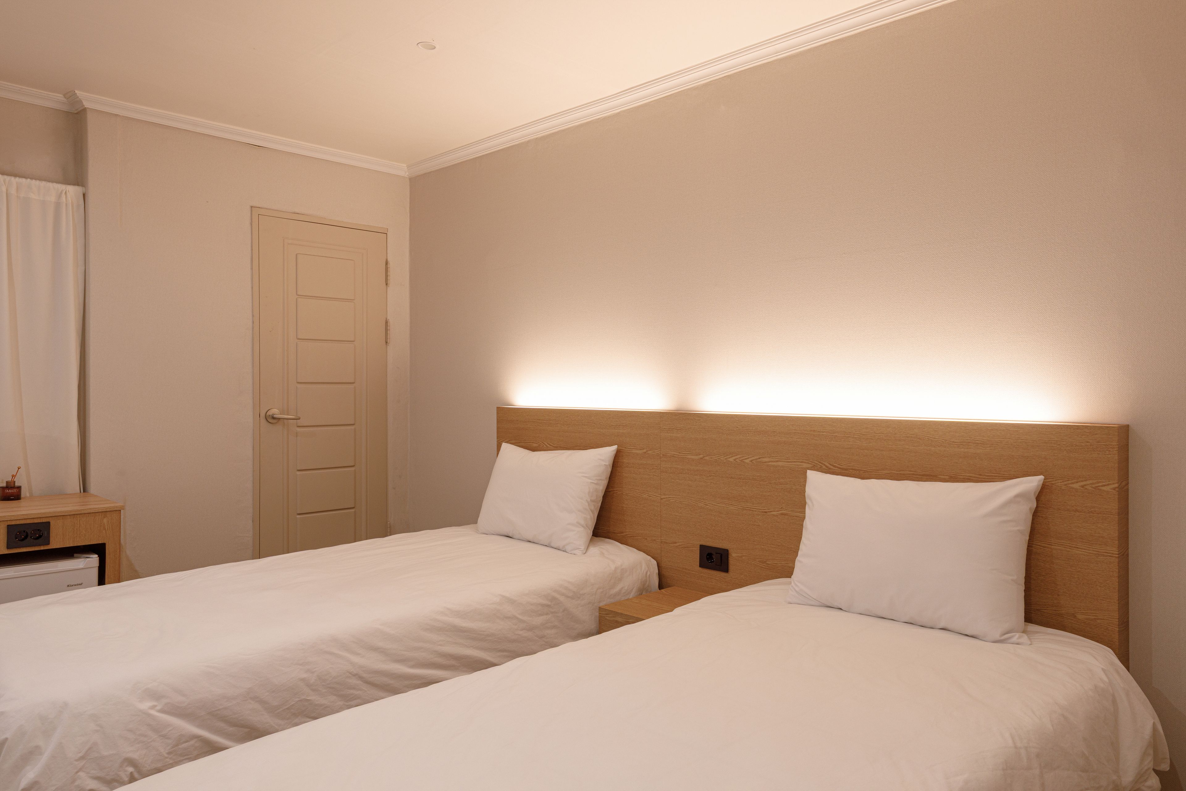 Quarto de hotel com duas camas, cabeceira de madeira, mesa de cabeceira e uma porta.