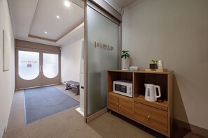 Quarto de hotel com porta deslizante, micro-ondas, chaleira, mesa pequena e planta em vaso.