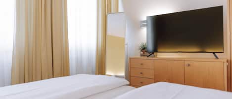 Quarto de hotel com cama, televisão sobre um móvel de madeira e uma janela grande com cortinas.