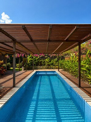 Piscina retangular com deck de madeira ao redor, sob uma pérgula de madeira.