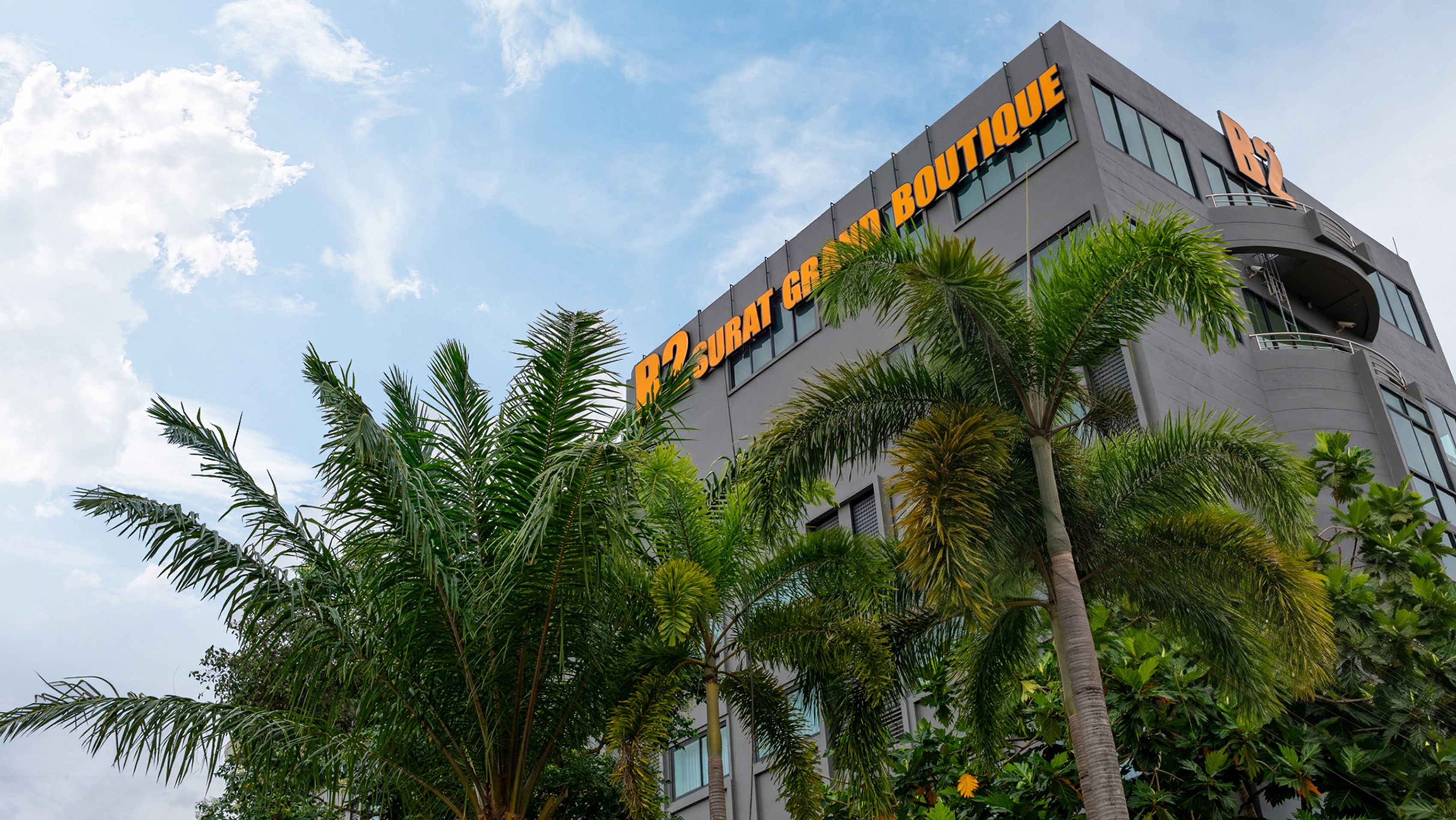 Um edifício com uma placa indicando 'R2 Surat Boutique' e cercado por palmeiras.