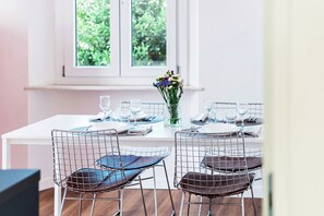 Uma sala de jantar com uma mesa branca posta para quatro pessoas, rodeada por cadeiras de estrutura metálica, e um vaso com flores sobre a mesa.