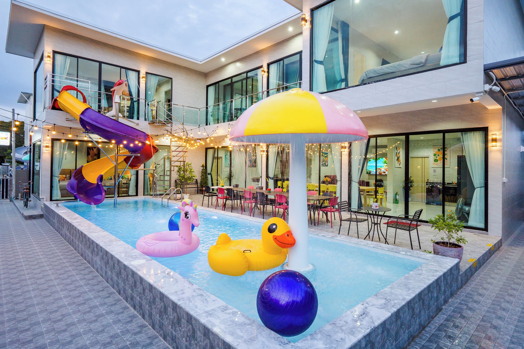 Uma área de piscina com brinquedos infláveis e um escorregador, cercada por assentos externos e um edifício com grandes janelas.
