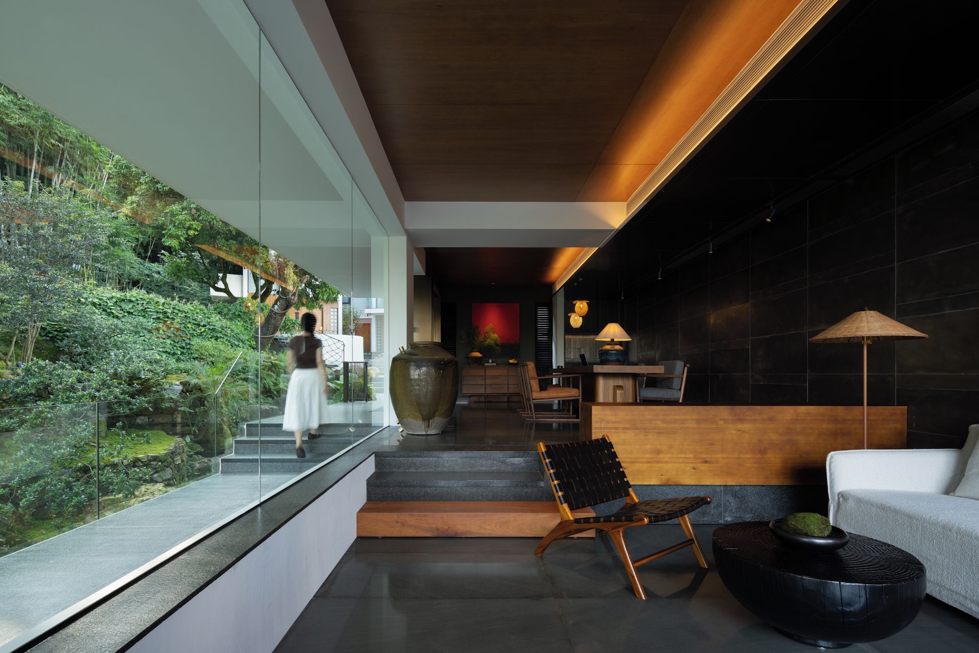 Interior moderno com parede de vidro dando para um jardim, um balcão de recepção e uma área de estar com abajur.