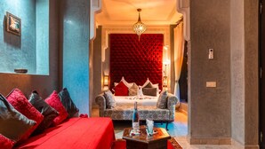 Quarto de hotel com carpete vermelho, cama com roupa de cama branca, mesa de cabeceira com uma garrafa e parede vermelha com padrão texturizado.