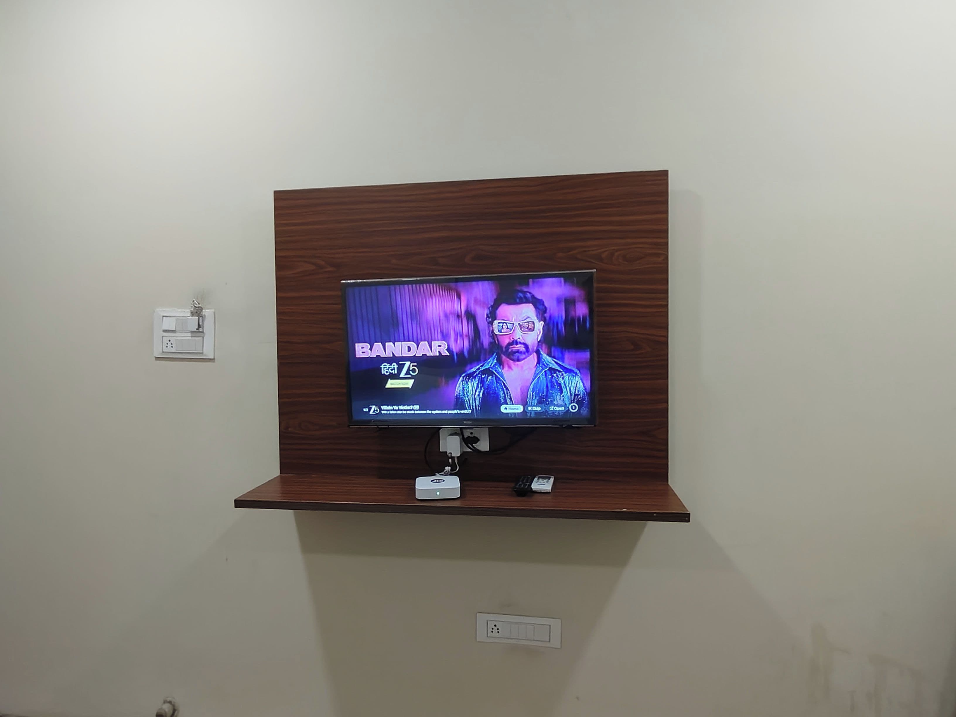 Televisão fixada numa parede de madeira com um comando a funcionar num suporte abaixo.