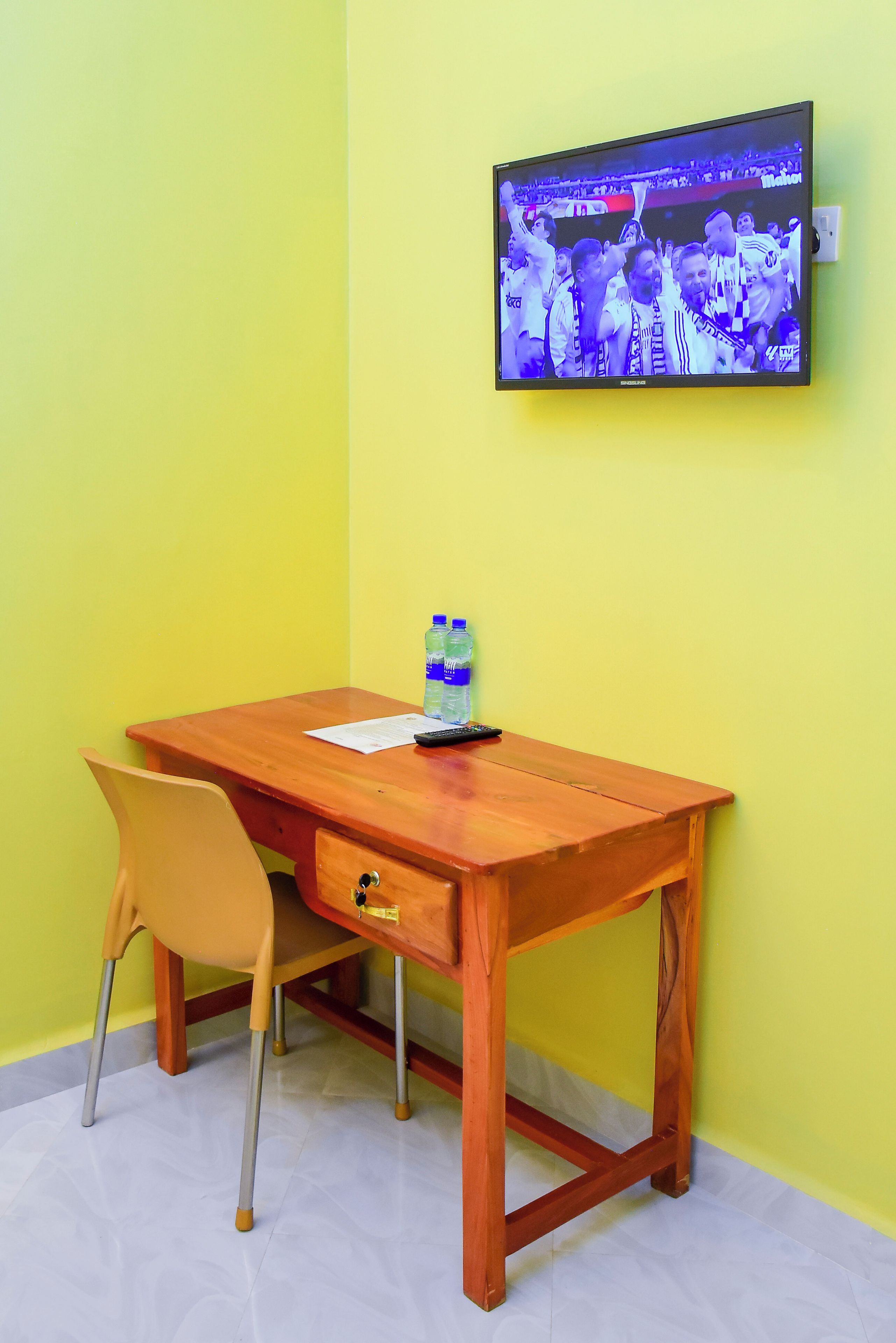 Uma mesa de madeira com uma cadeira, uma televisão presa na parede e uma garrafa de água sobre a mesa.