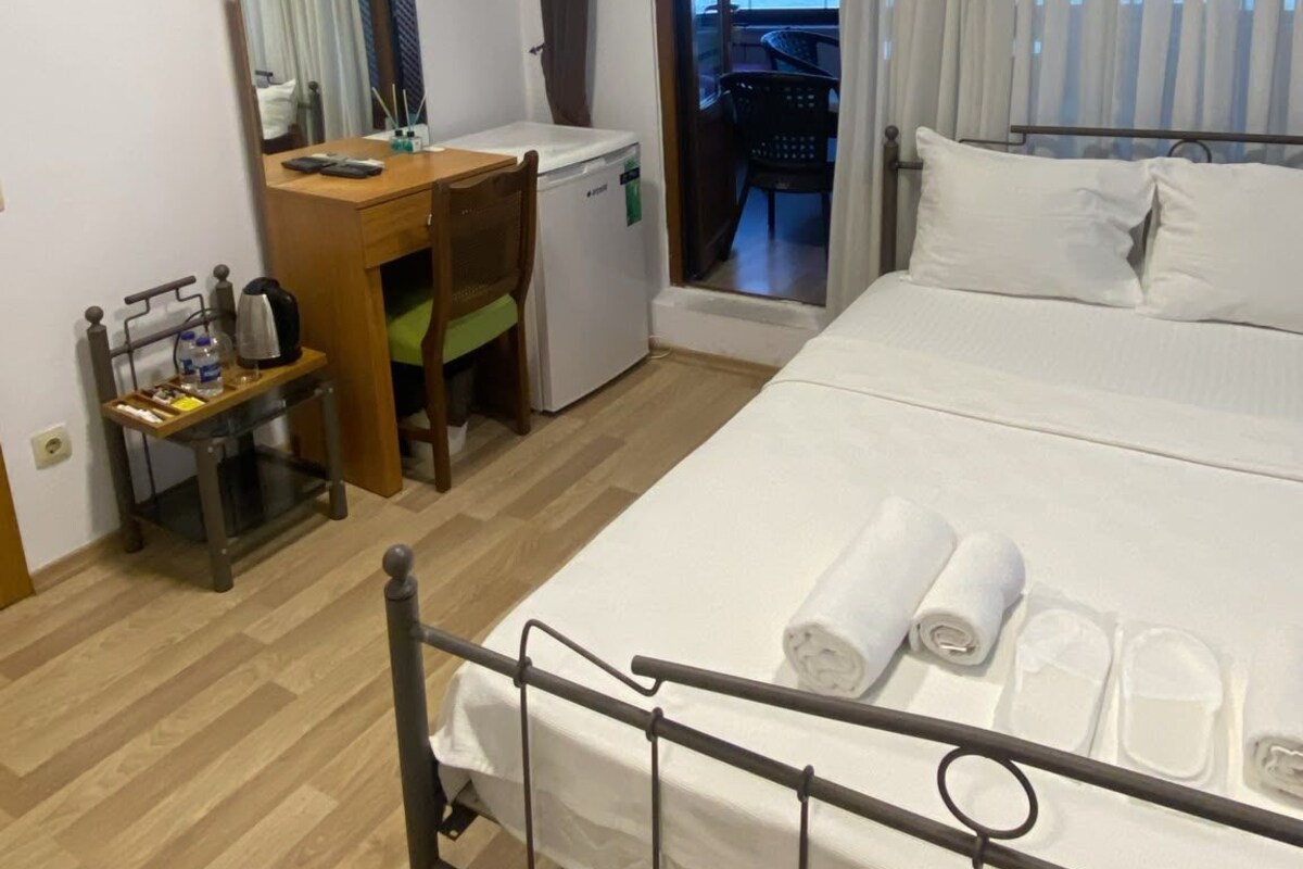 Quarto de hotel com cama, escrivaninha, cadeira, pequeno frigorífico, televisão e varanda com vista.