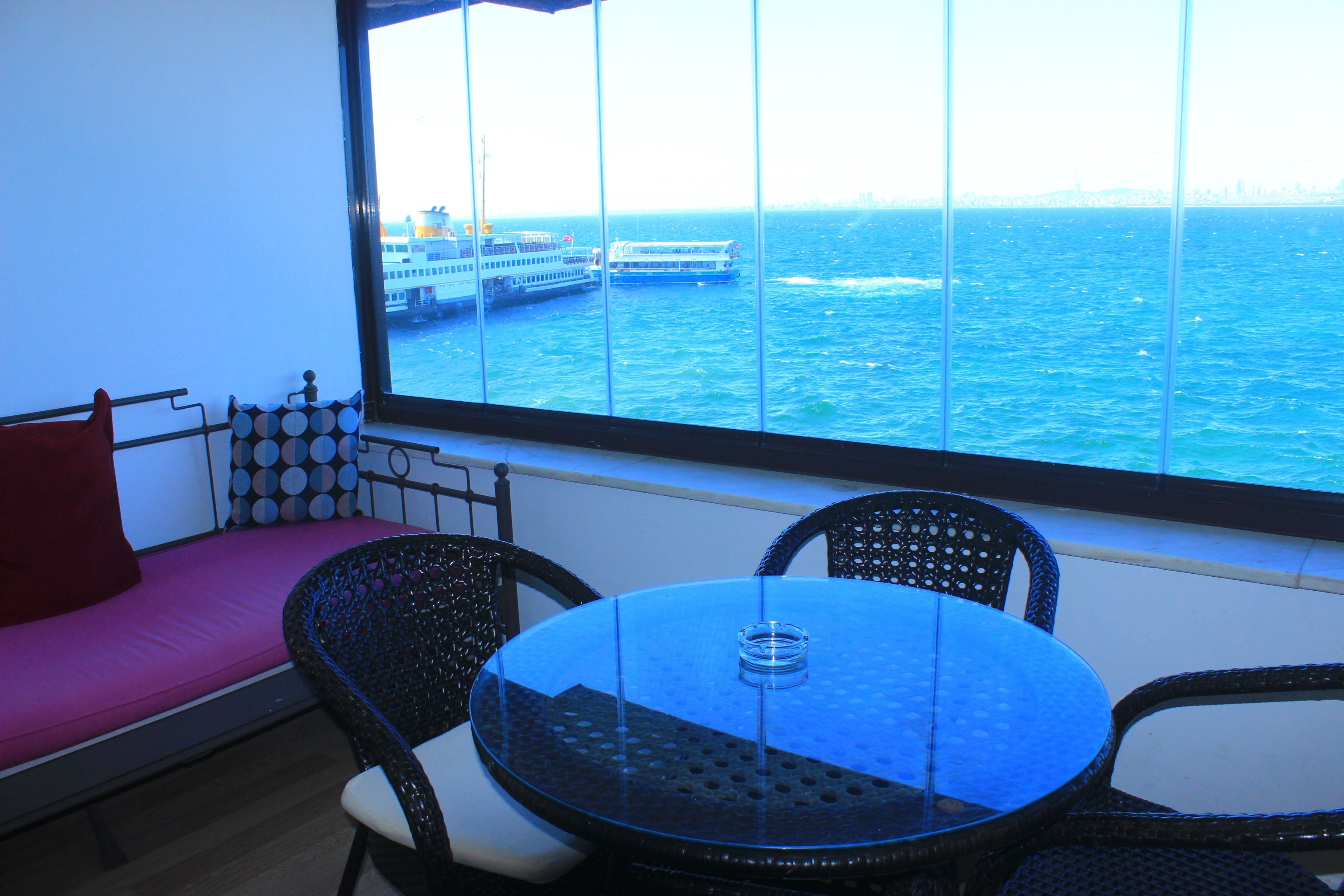 Varanda com vista para o mar e um barco, mesa redonda com um copo e cadeiras de vime.