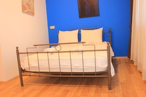Uma cama individual com estrutura de metal e roupa de cama branca, diante de uma parede azul com um quadro em cima.