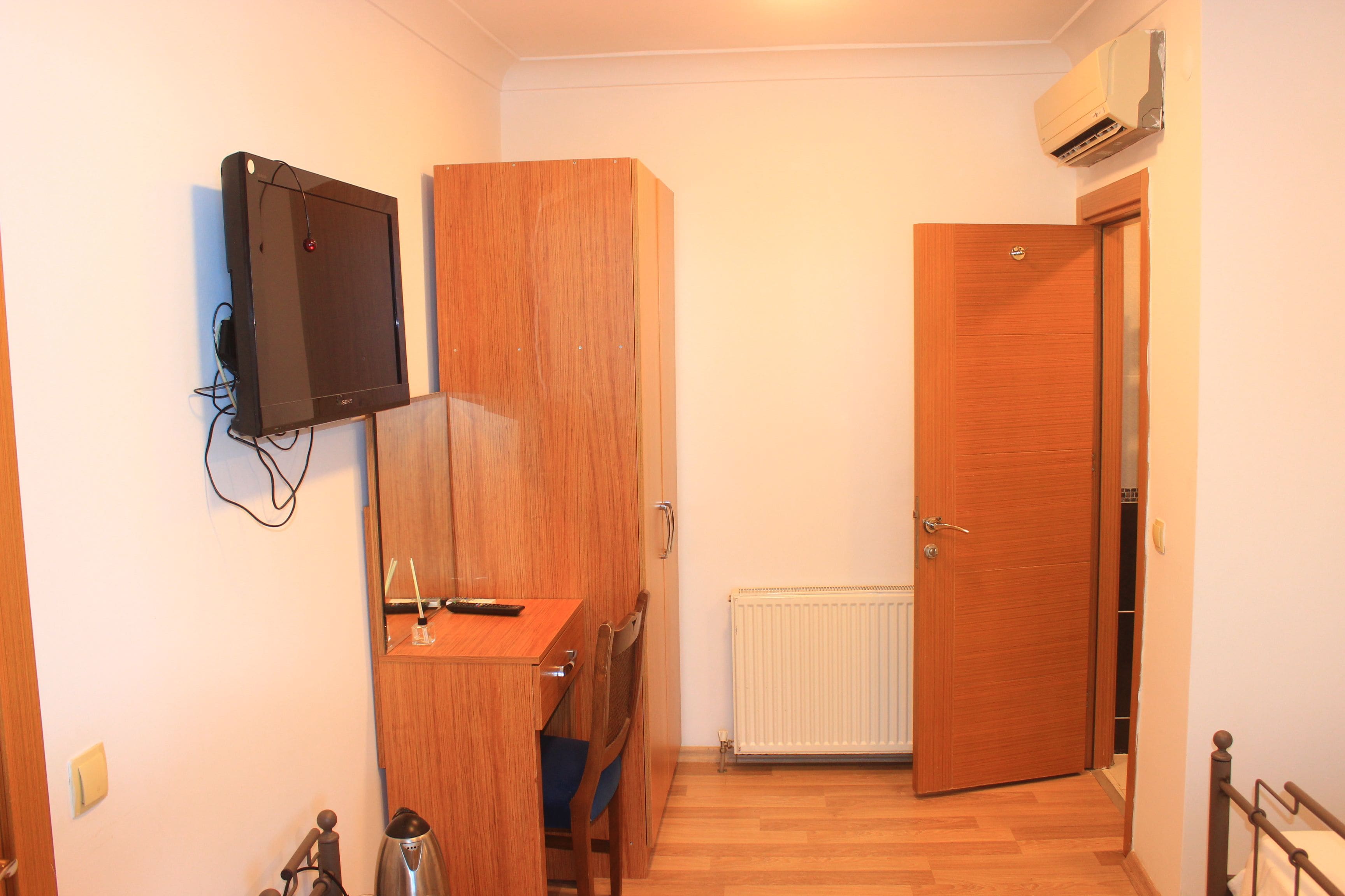 Quarto pequeno com guarda-roupa de madeira, escrivaninha com cadeira, radiador e televisão na parede.