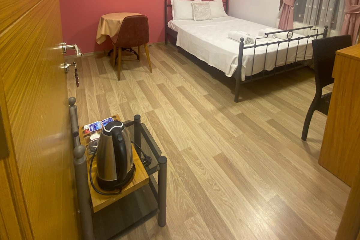 Quarto de hotel com cama, mesa pequena, cadeira, chaleira e janela com cortinas.