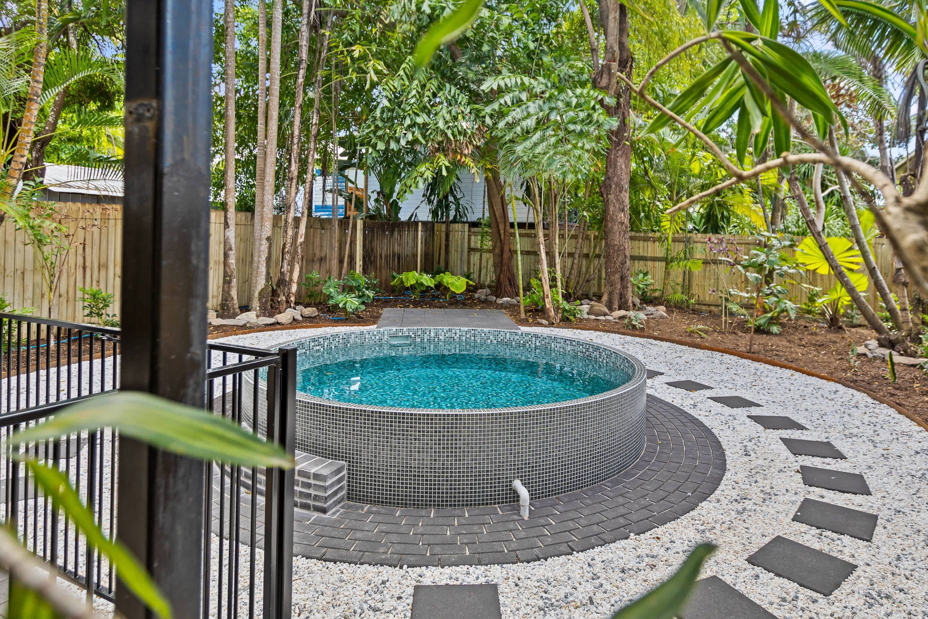 Uma banheira de hidromassagem circular rodeada por uma borda de pedrinhas, com uma cerca de madeira e plantas tropicais ao fundo.