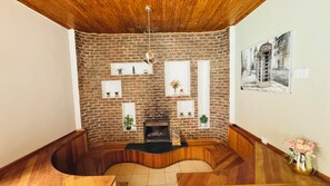 Quarto com parede de tijolos, teto de madeira e lareira. Há prateleiras com plantas e um quadro na parede.