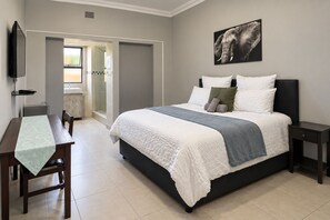 Quarto com cama, mesa de cabeceira, escrivaninha e uma janela grande.