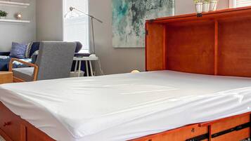 Quarto com estrutura de cama de madeira, colchão branco, escrivaninha com cadeira e janela com persianas.