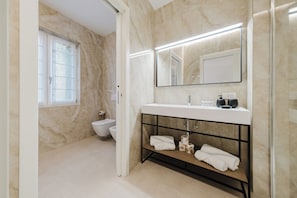 Uma casa de banho moderna com parede de mármore, sanita, lavatório com espelho e prateleira com toalhas.