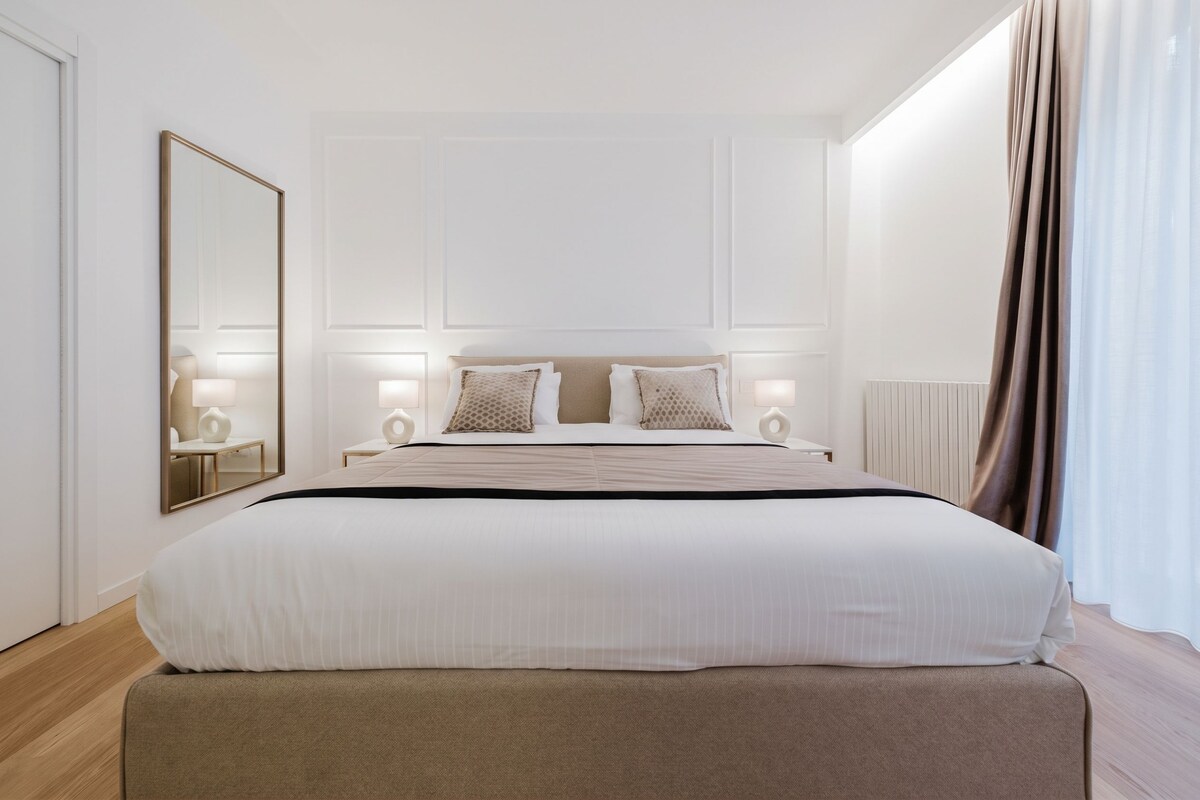 Quarto de hotel com uma cama grande, mesas de cabeceira, um espelho e uma janela com cortinas.