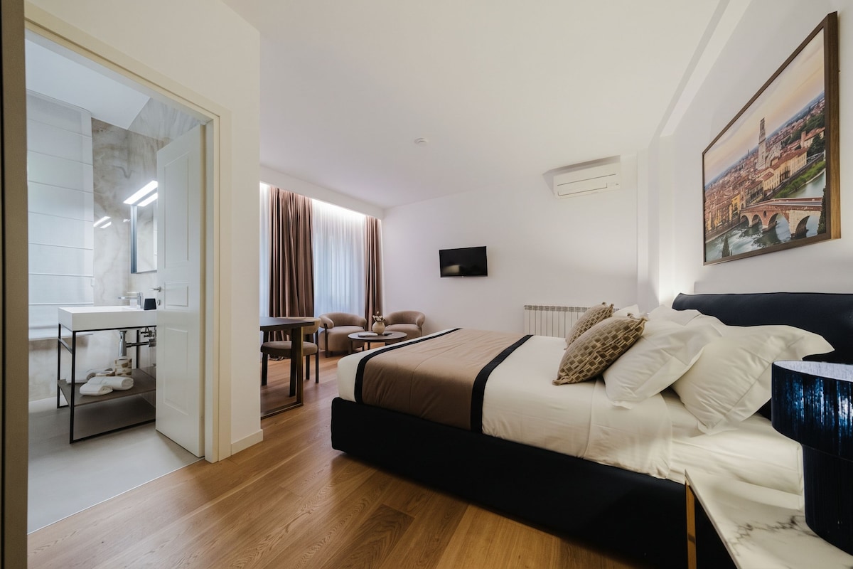 Quarto de hotel moderno com cama, escrivaninha, cadeira, televisão e um quadro na parede.