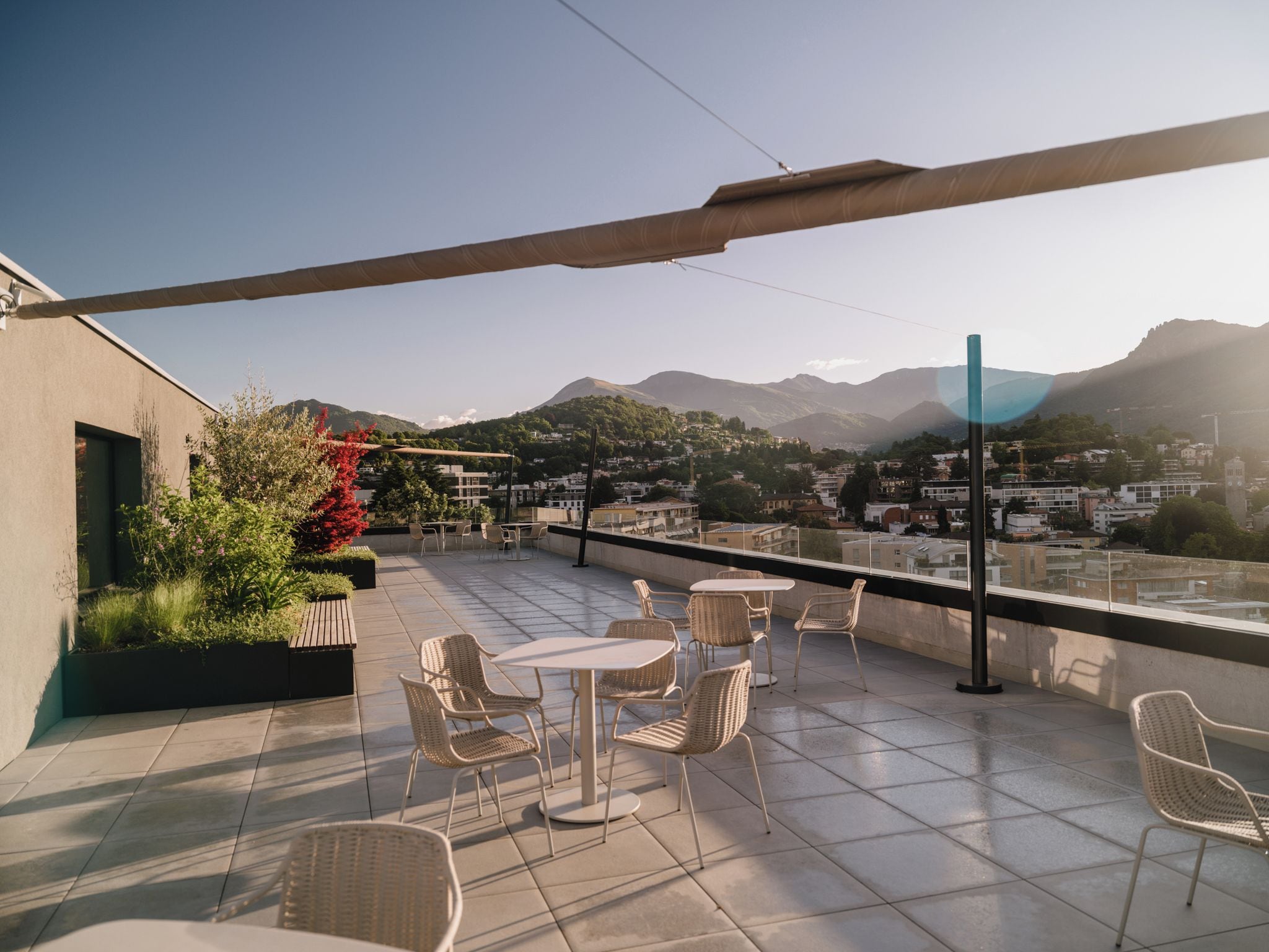 Um terraço com mesas ao ar livre, vista para uma cidade e montanhas ao fundo.