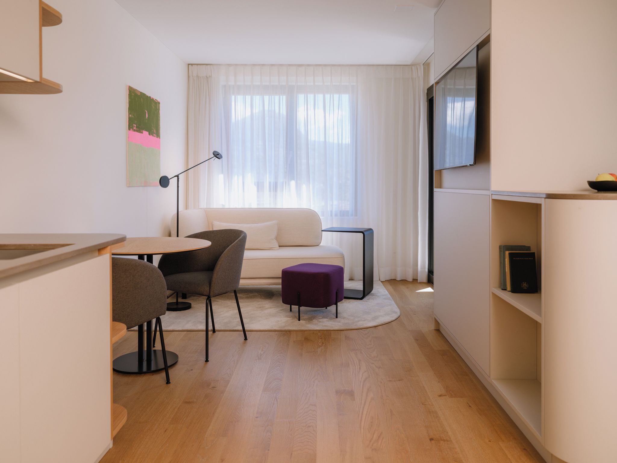 Uma sala de estar moderna com um sofá, duas poltronas, uma mesinha, um banquinho roxo, um abajur e uma TV de tela plana fixada na parede.