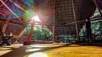 Um espaço interno com telhado de palha, vigas de madeira e uma fonte de luz central.