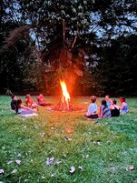 Um grupo de pessoas sentadas ao redor de uma fogueira em uma área gramada, com árvores ao fundo.