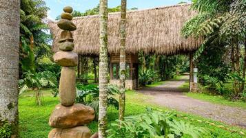 Uma construção com telhado de palha rodeada por vegetação exuberante e uma escultura de pedra equilibrada.