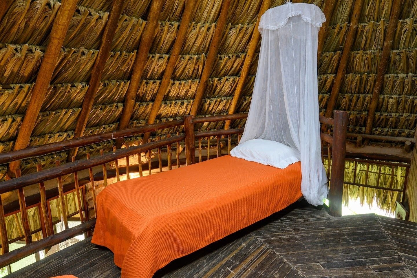 Uma cama de solteiro com colcha laranja e almofadas brancas, sob uma estrutura de telhado de palha.