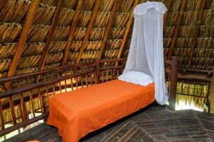 Uma cama de solteiro com colcha laranja e almofadas brancas, sob uma estrutura de telhado de palha.