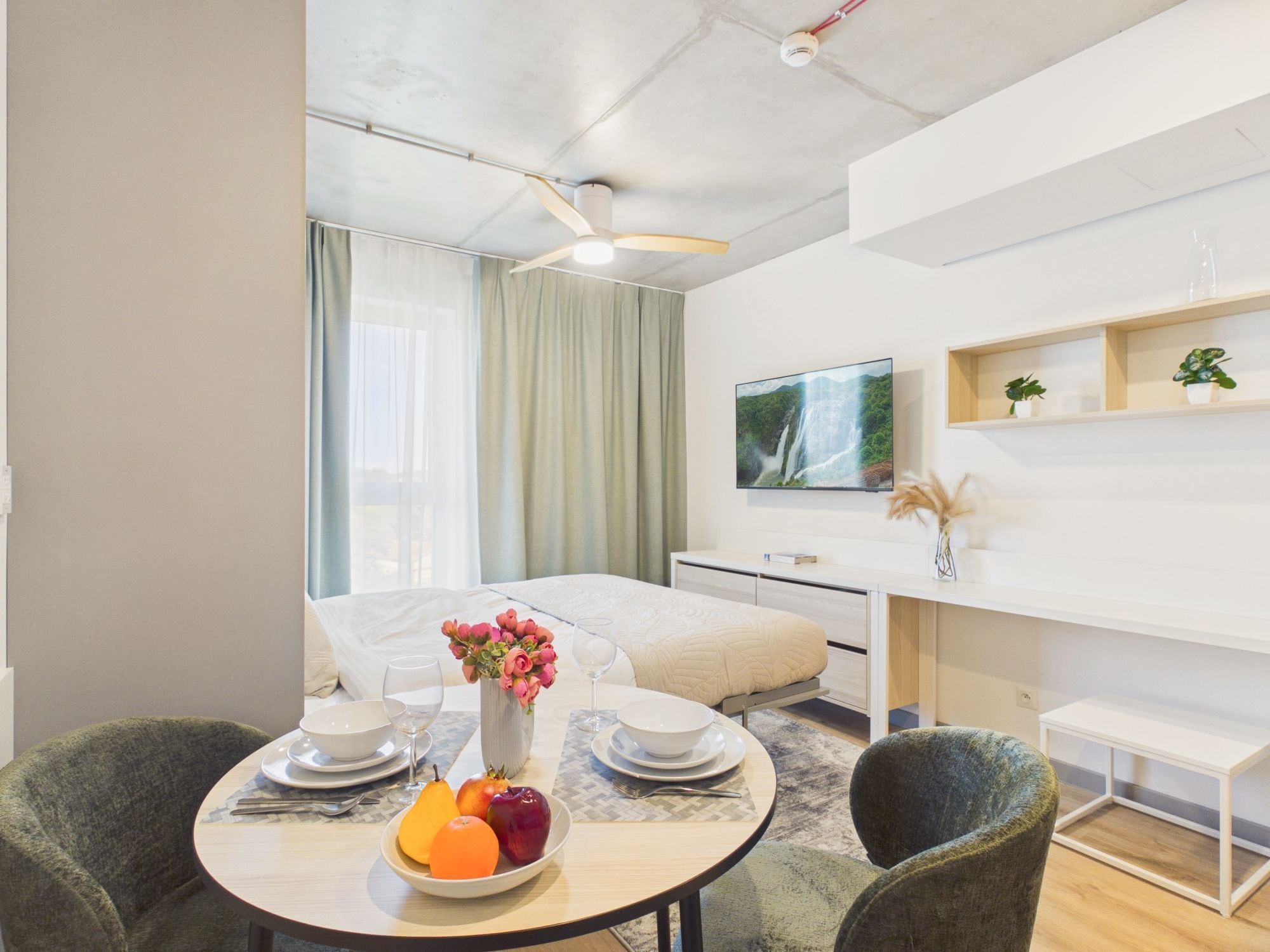 Quarto moderno com cama, mesa de cabeceira, televisão e um espaço de refeições com frutas e flores.