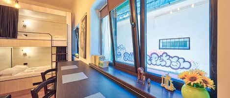 Quarto de hotel com beliche, mesa de jantar e uma grande janela com obras de arte.