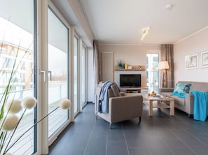 Uma sala de estar moderna com sofá, poltrona e mesa de centro, televisão e uma janela grande com vista.
