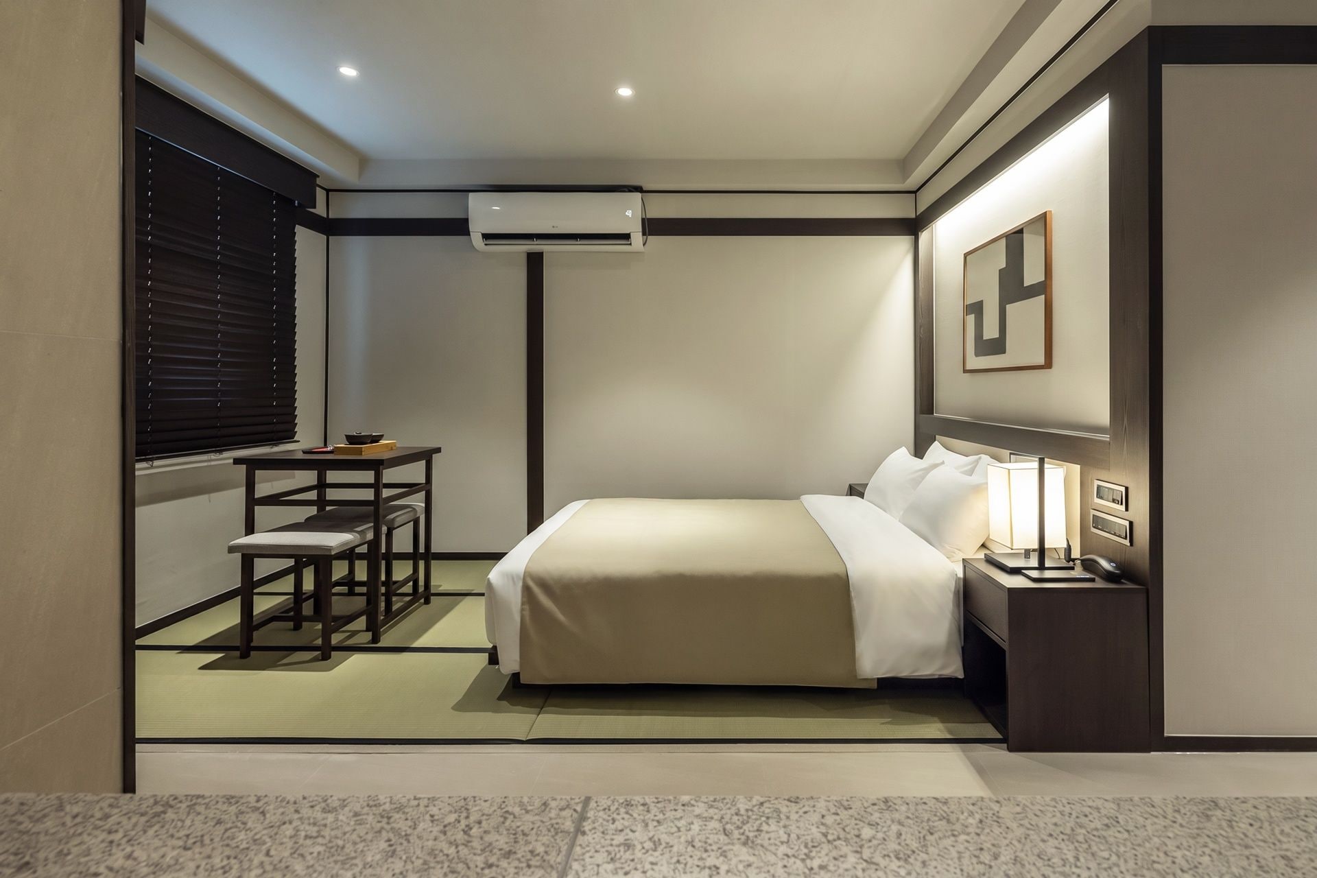 Quarto de hotel com cama, mesa de cabeceira, luminária, ar condicionado e uma pequena mesa com cadeira.
