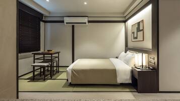 Quarto de hotel com cama, mesa de cabeceira, luminária, ar condicionado e uma pequena mesa com cadeira.