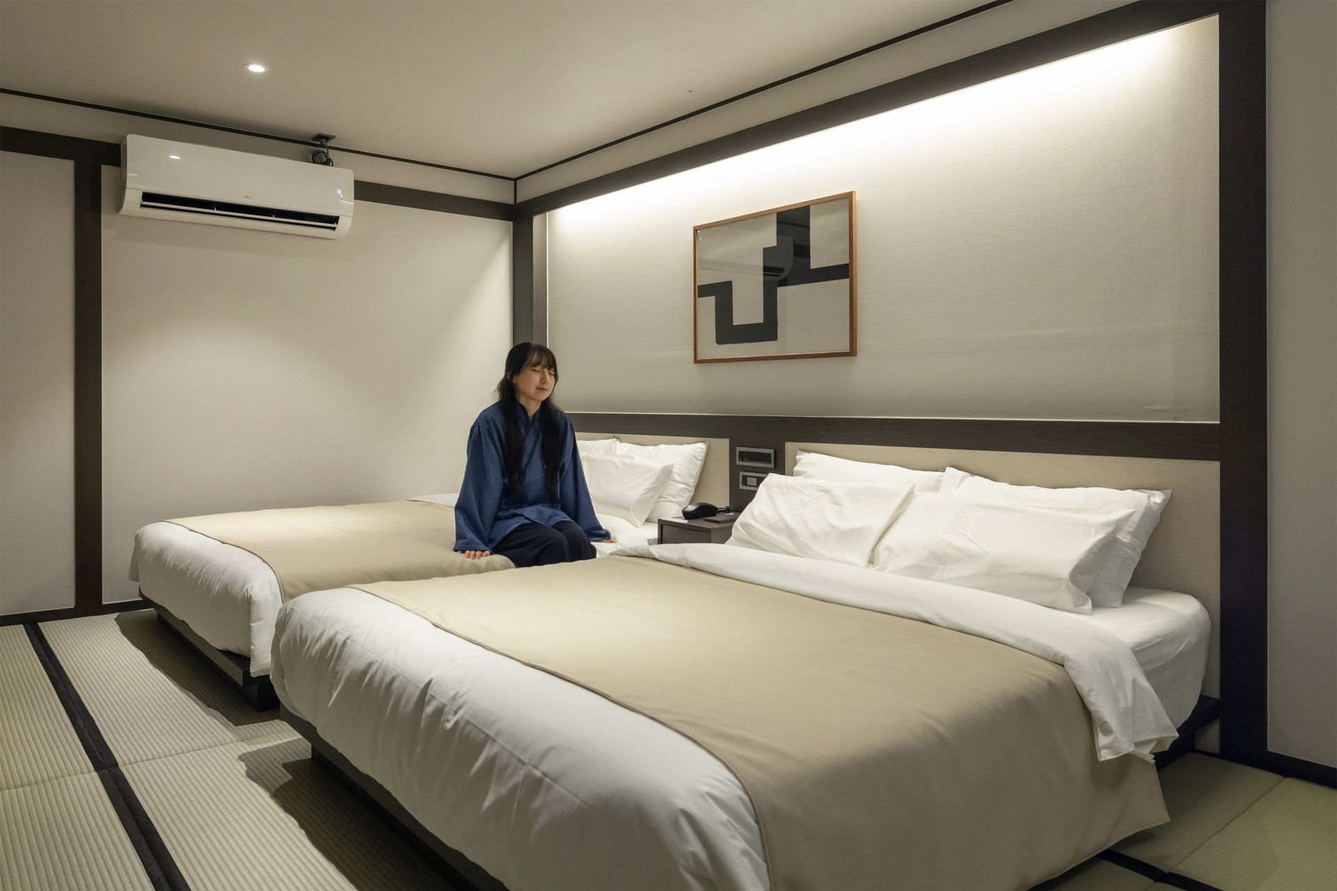 Quarto de hotel com duas camas, ar condicionado fixado na parede, mesa de cabeceira com abajur e uma obra de arte abstrata emoldurada na parede.