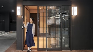 Uma pessoa em frente à entrada de um prédio, com porta de vidro, placa com a marca do hotel e interior moderno com iluminação visível.