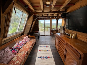 Um quarto de sótão acolhedor com teto de madeira, um sofá com almofadas coloridas, uma mesa de jantar e uma televisão.