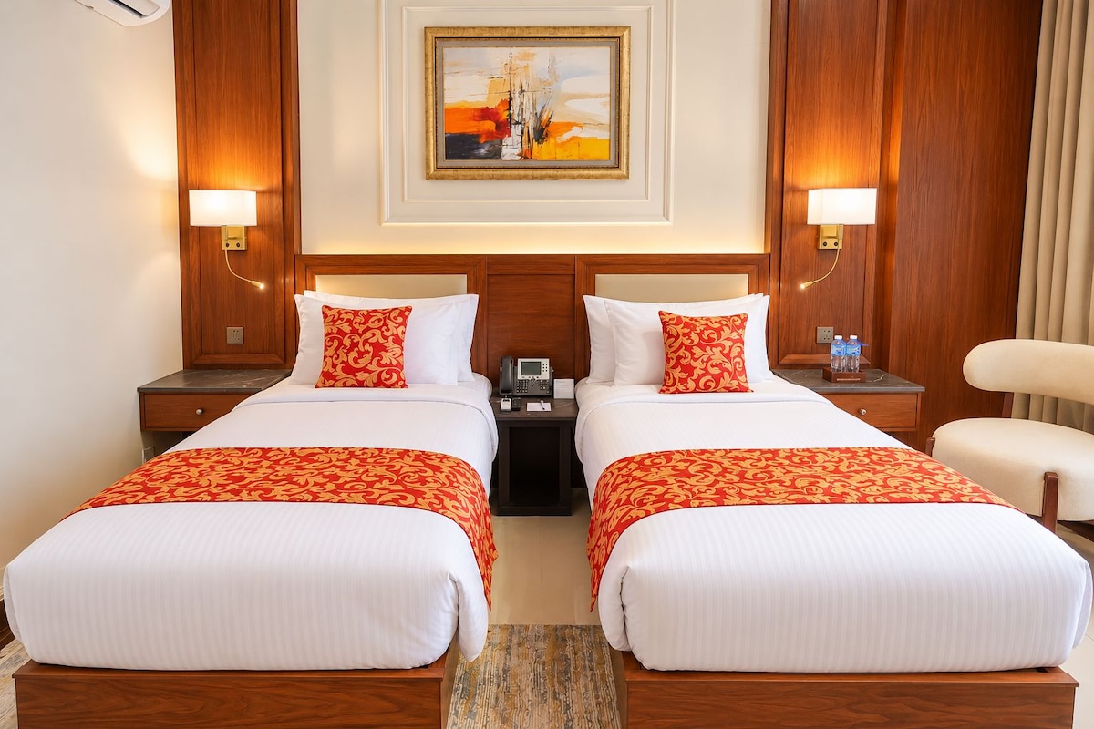 Quarto de hotel com duas camas, um quadro acima das camas, abajures, telefone e uma cadeira.