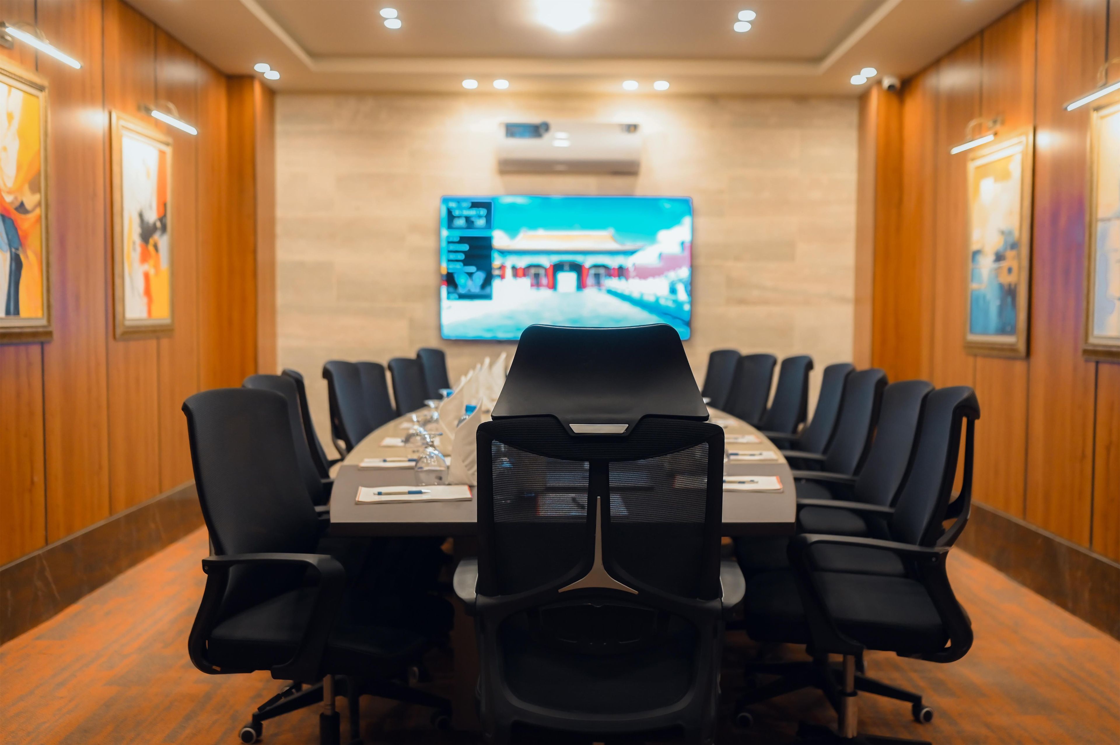 Uma sala de conferências com uma mesa comprida, cadeiras pretas e uma tela grande na frente.