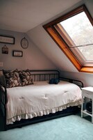 Quarto com teto inclinado, claraboia, cama com colcha branca, mesa de cabeceira e relógio de parede.
