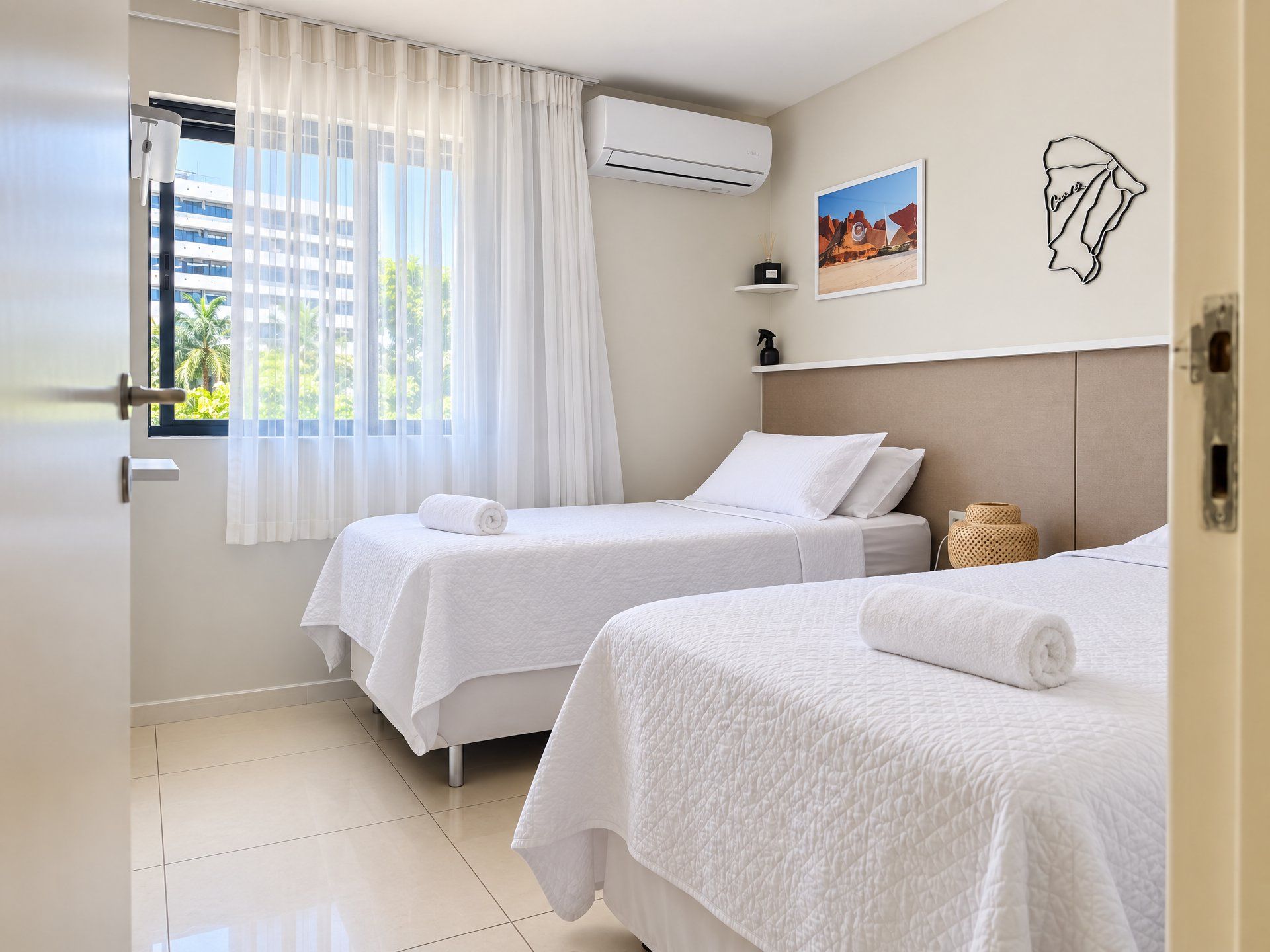 Quarto de hotel com duas camas, janela com cortinas, ar condicionado, prateleira pequena com uma garrafa e quadro emoldurado na parede.