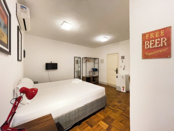 Quarto de hotel com cama, luminária de mesa vermelha, televisão, guarda-roupa e um aviso que diz 'CERVEJA GRÁTIS Amanhã'.